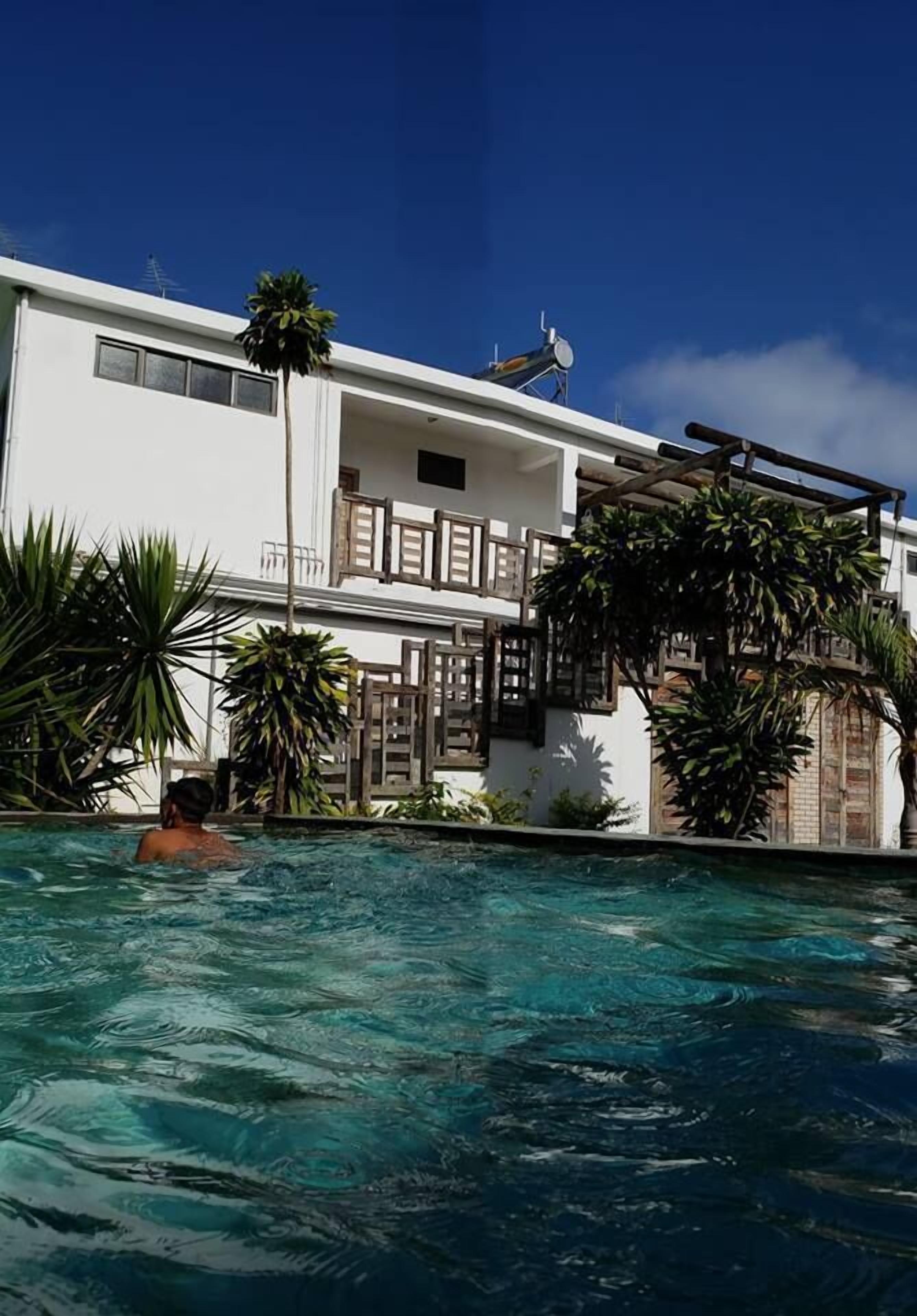 Uma área de piscina com um edifício branco ao fundo, cercada por plantas tropicais.