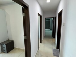 Um corredor com uma porta de madeira escura à esquerda, piso de azulejos claros e um pequeno armário com gavetas.