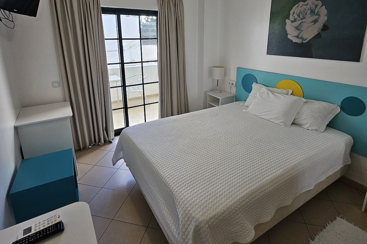 Quarto de hotel com uma cama, televisão, janela com cortinas, um quadro e uma pequena mesa com um controle remoto.
