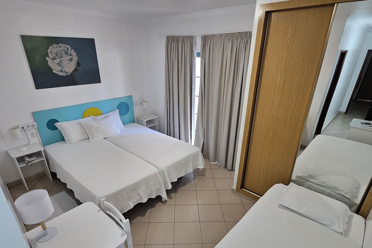 Quarto de hotel com duas camas, uma escrivaninha, uma cadeira, uma mesa de cabeceira, um abajur, um quadro e uma janela com cortinas.