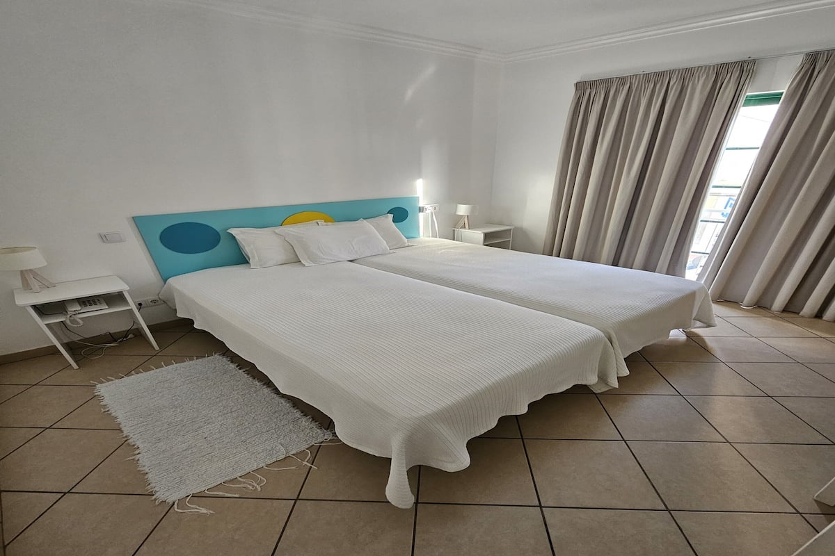 Uma cama de casal com cabeceira azul, roupa de cama branca, uma pequena mesa de cabeceira com abajur e uma janela com cortinas.