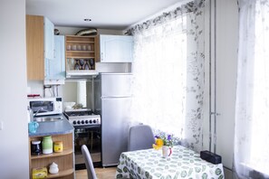 Uma cozinha compacta com fogão, geladeira e micro-ondas. Há uma mesa de jantar com toalha de mesa estampada e uma cadeira.