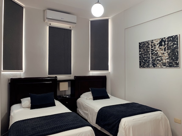 Quarto de hotel com duas camas, um ventilador de teto, ar condicionado, mesa de cabeceira, luminária e uma obra de arte na parede.