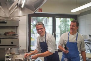 Dois chefs em uma cozinha com eletrodomésticos de aço inoxidável, uma janela com vista para árvores e um alarme de incêndio na parede.