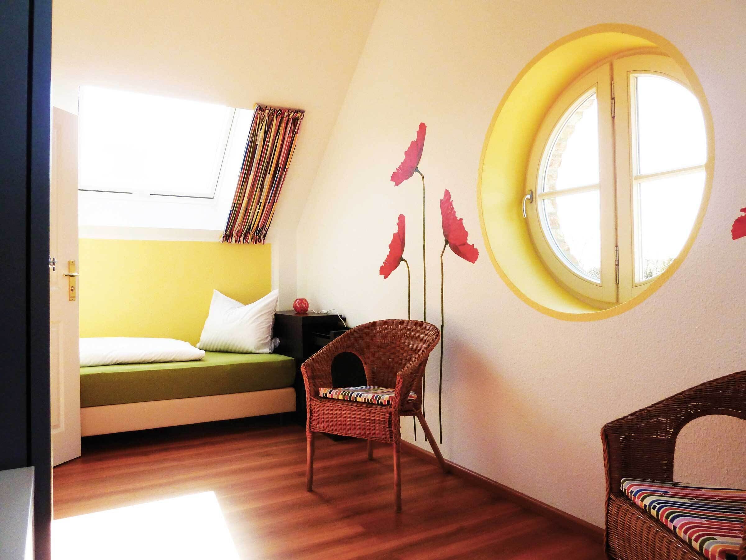 Quarto com cama, cadeira, mesa pequena, janela e parede com janela circular.