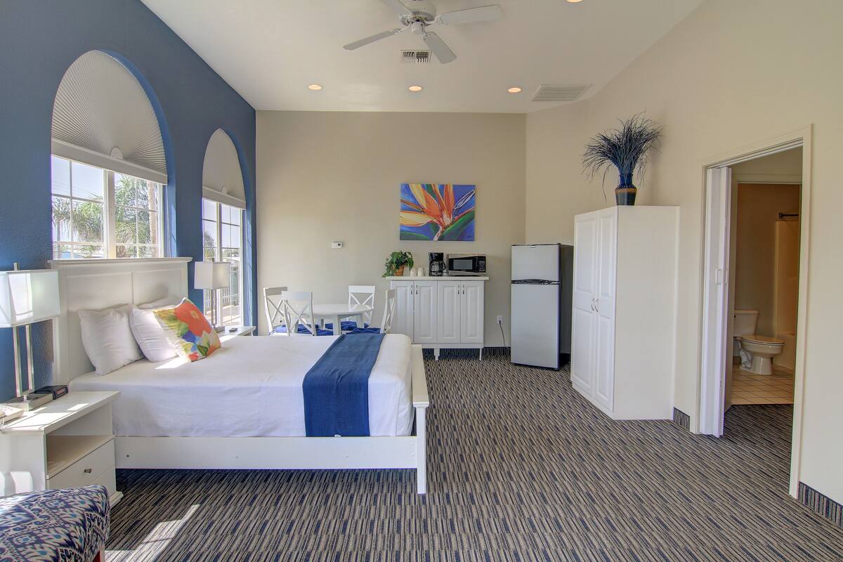 Quarto de hotel com cama, escrivaninha, cadeira, cozinha compacta e banheiro.