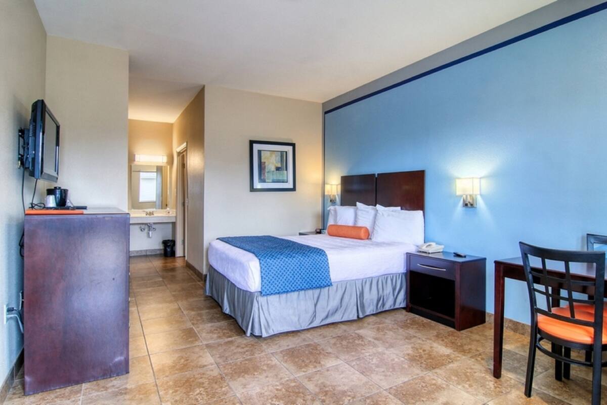 Quarto de hotel com cama, mesa de cabeceira, cadeira, escrivaninha, televisão e banheiro ao fundo.