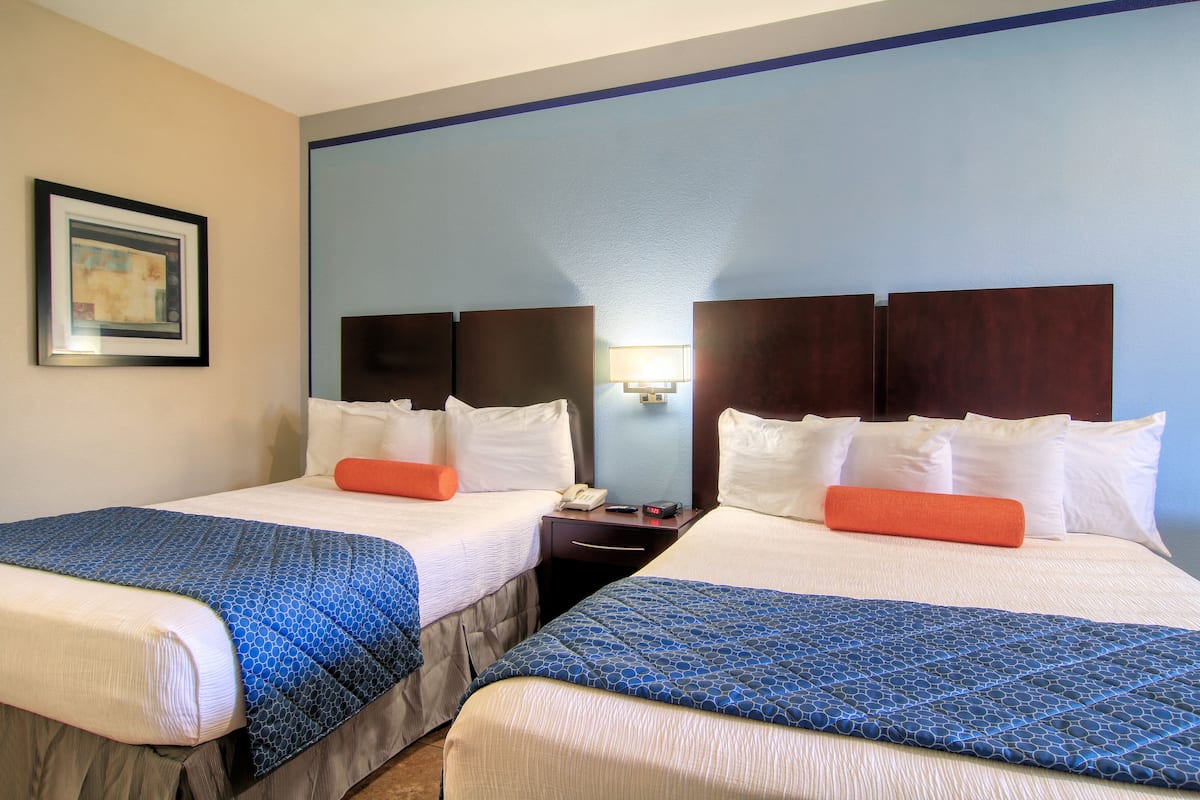 Duas camas de casal com roupa de cama azul e branca, uma mesa de cabeceira com telefone e controle remoto, e um quadro pendurado na parede.