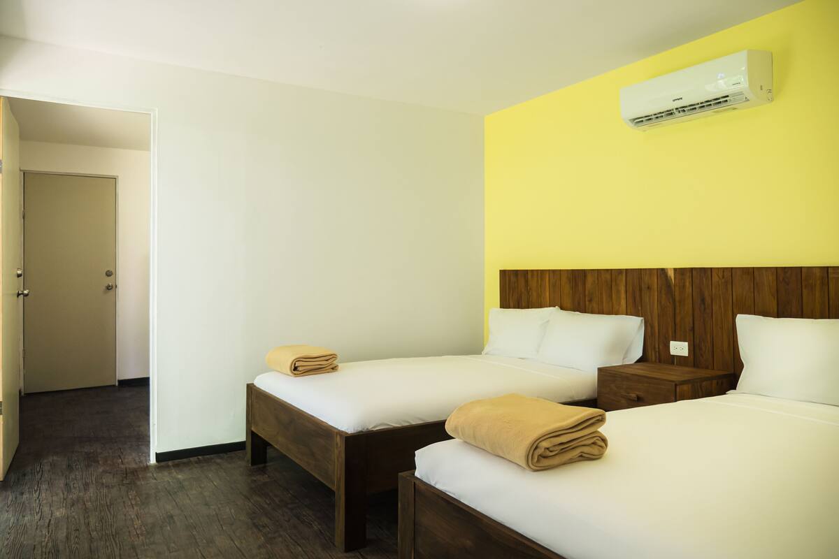 Quarto de hotel com duas camas, cabeceiras de madeira, chão de madeira, porta branca e um aparelho de ar condicionado na parede.