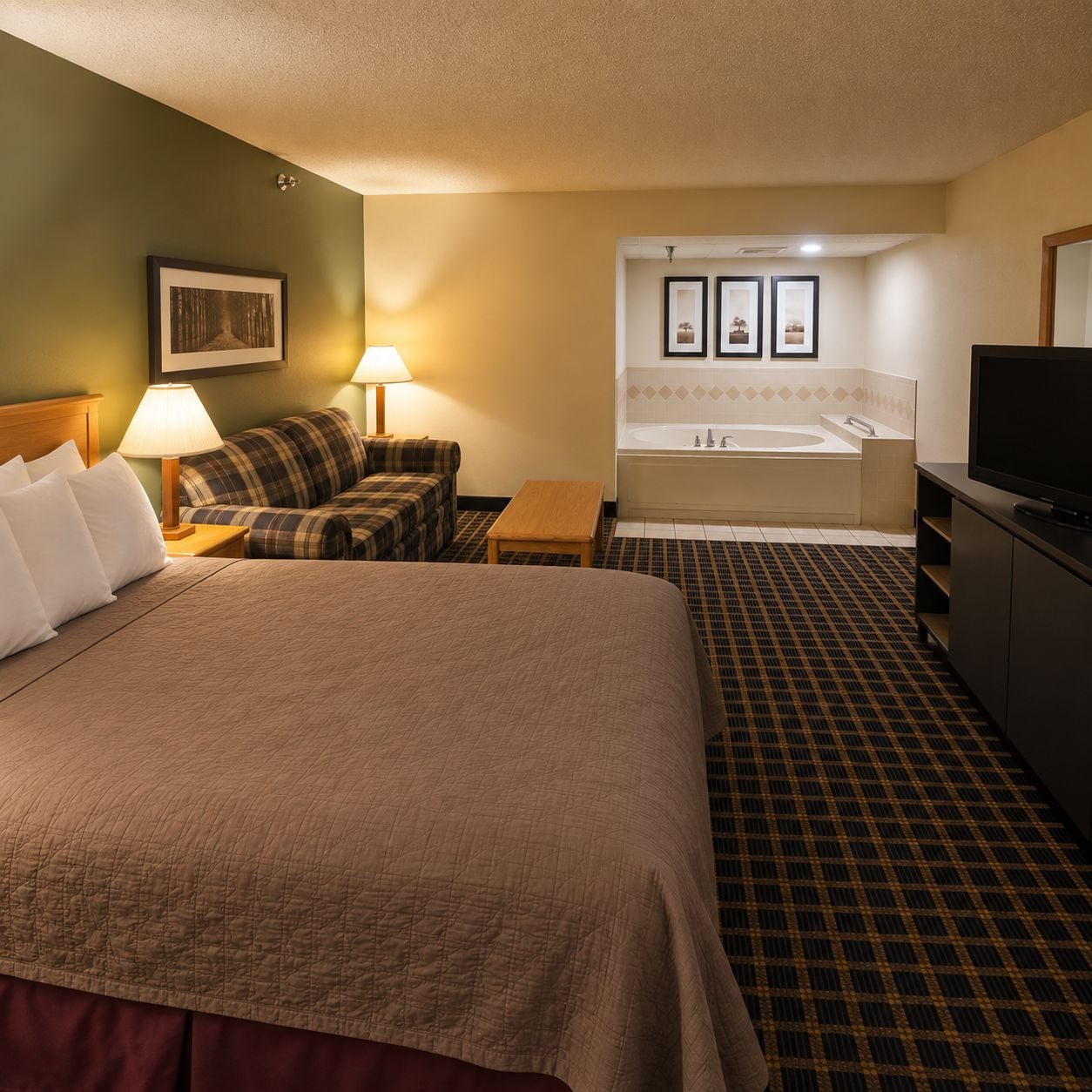 Quarto de hotel com uma cama grande, um sofá, uma mesinha, uma televisão e banheiro com banheira e vaso sanitário.