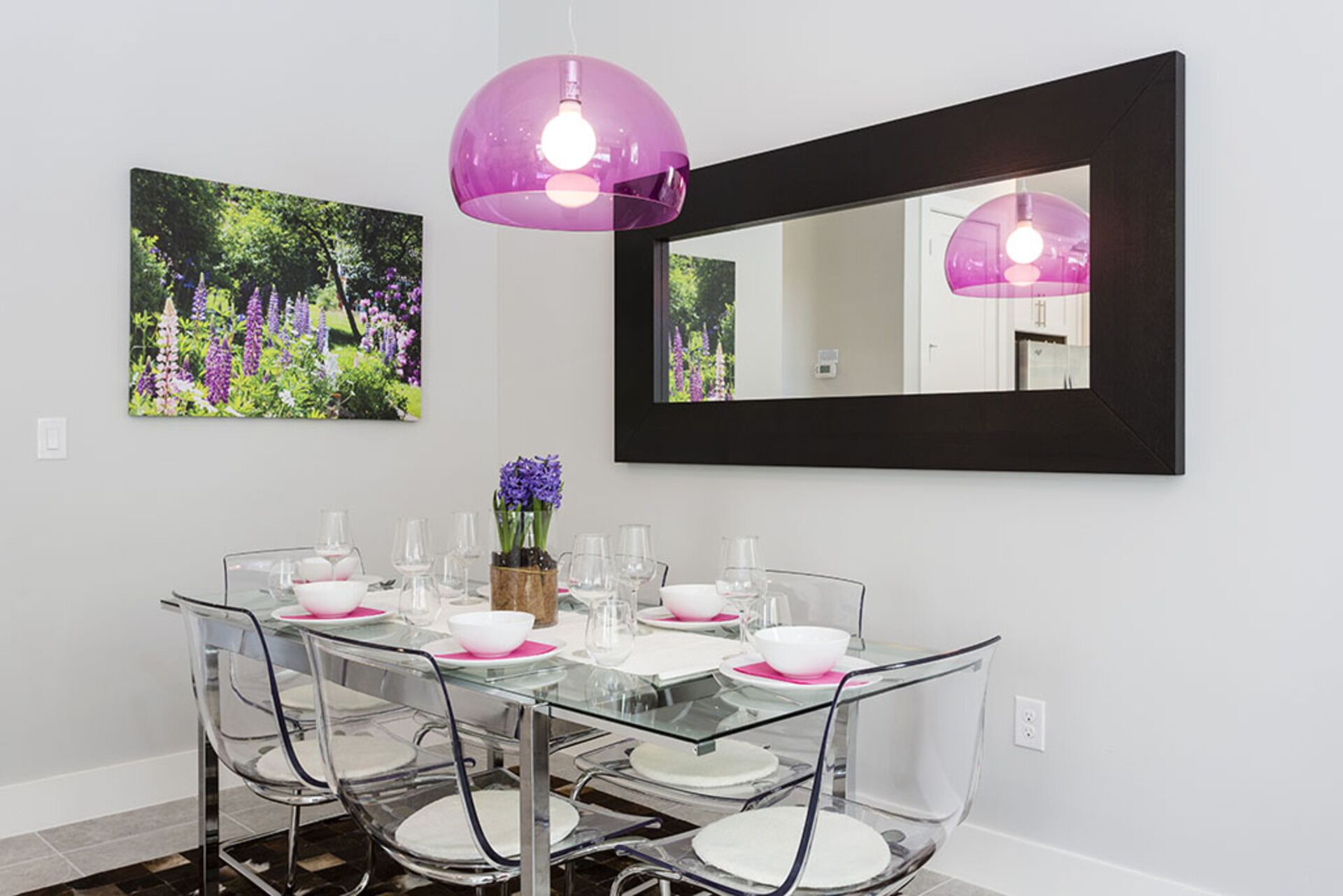 Uma área de refeições com mesa de vidro posta para quatro pessoas, cadeiras transparentes, um vaso com flores roxas e um espelho grande na parede.