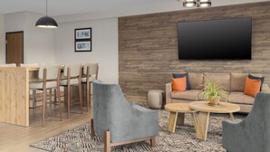 Uma sala de estar moderna com área de bar, poltronas estofadas em cinza, um sofá com almofadas laranja e uma televisão de tela plana grande fixada em uma parede de madeira.