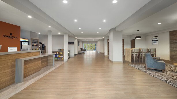 Uma área de recepção de hotel com balcão, sala de espera com cadeiras, espaço de refeições com mesas e cadeiras, e um corredor que leva a outros quartos.