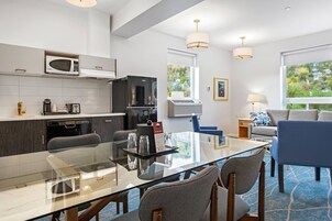 Quarto de hotel moderno com mesa de jantar de vidro, frigorífico preto, micro-ondas e área de estar com sofá e poltronas azuis.