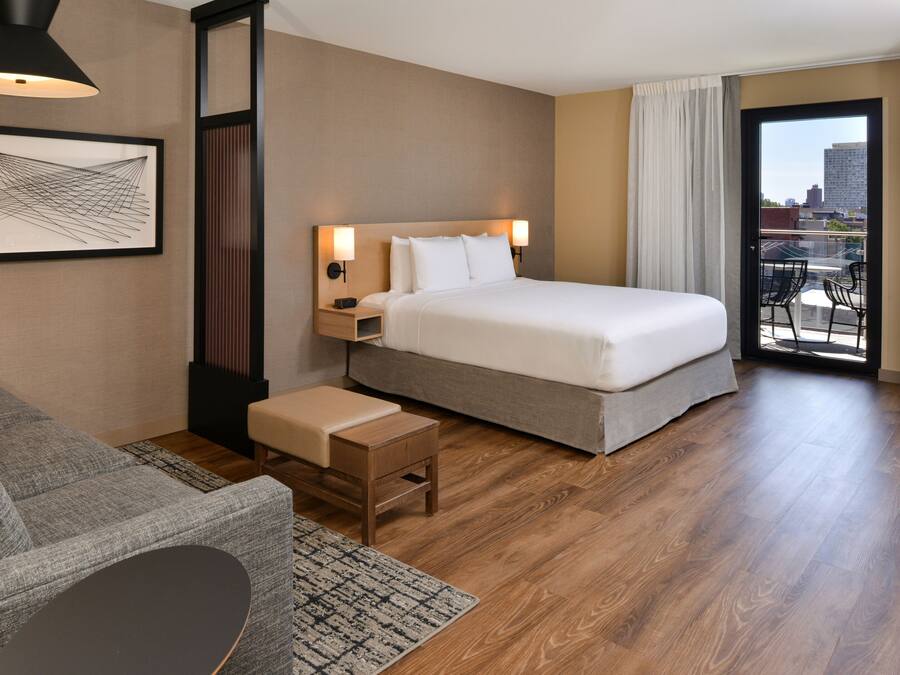 Quarto de hotel moderno com uma cama grande, um sofá, uma mesinha e uma varanda com vista para a cidade.