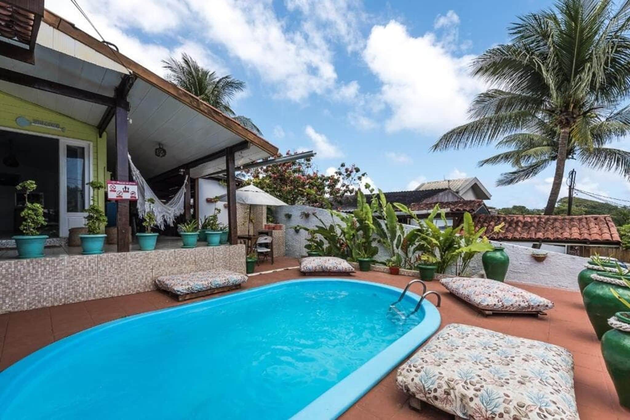 Uma piscina com espreguiçadeiras, plantas em vasos e um edifício com telhado de azulejos.