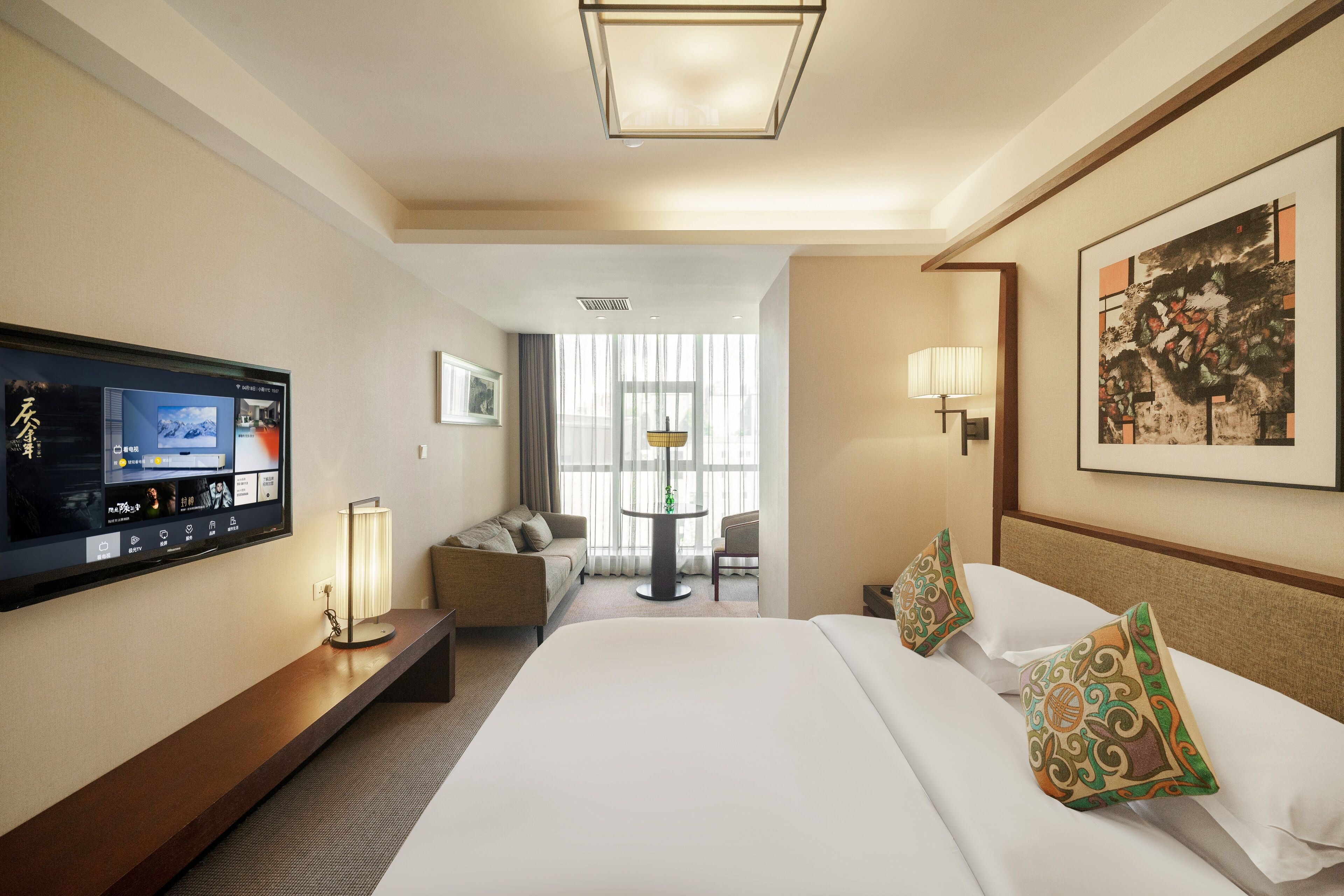 Quarto de hotel com uma cama grande, televisão de tela plana na parede, escrivaninha com luminária, sofá e janela com cortinas leves.