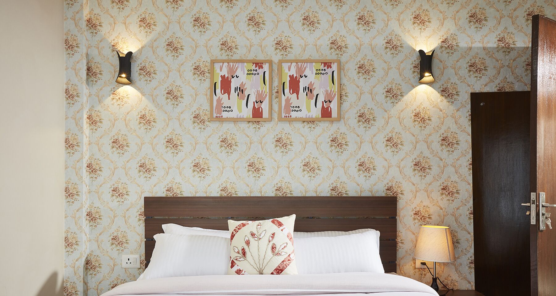 Quarto de hotel com papel de parede floral, cabeceira de madeira, duas luminárias fixadas na parede e cama com roupa de cama branca e um travesseiro decorativo.