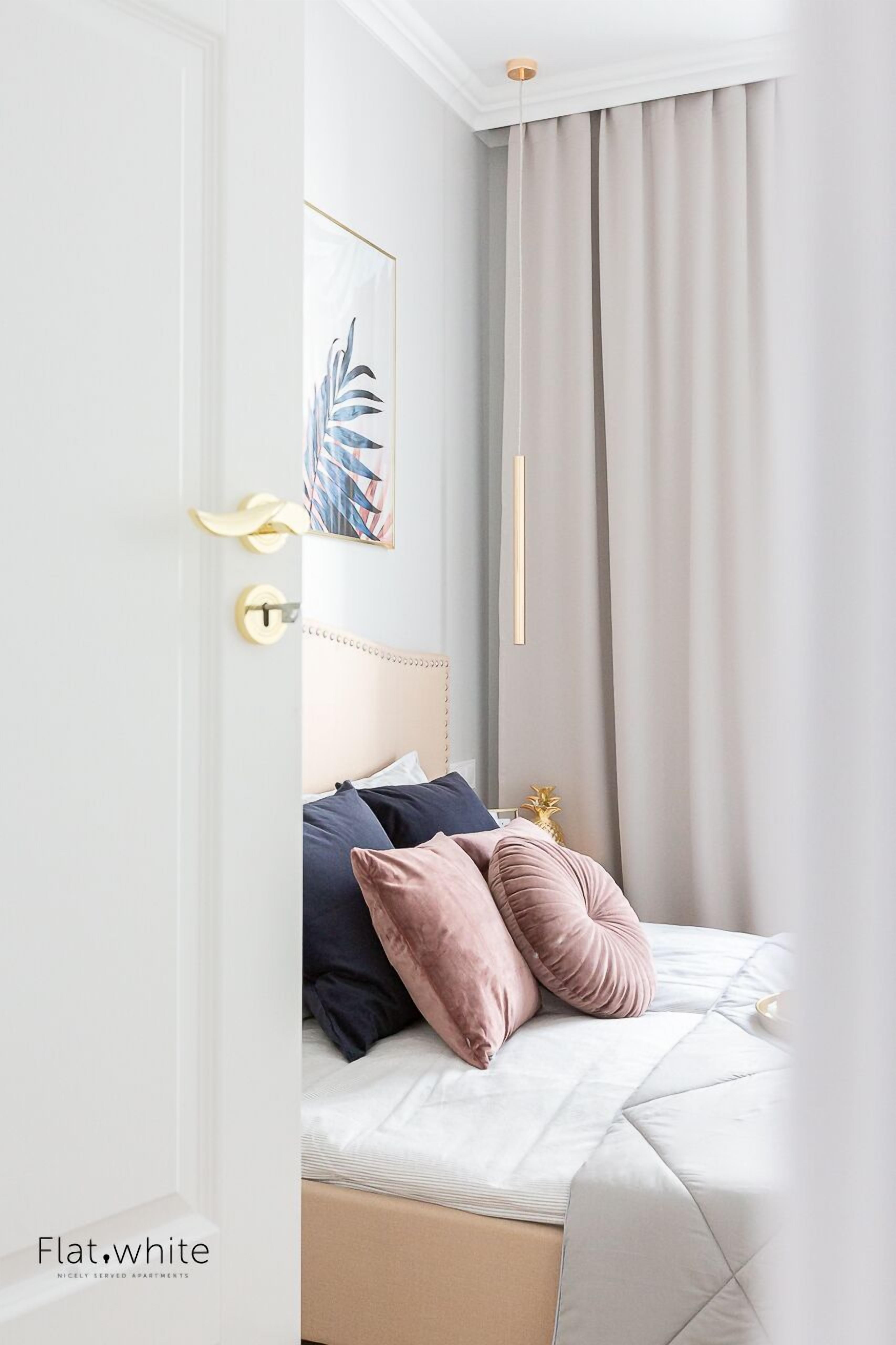 Quarto com cama, almofadas, cabeceira, obra de arte na parede, cortina e porta com luminária.
