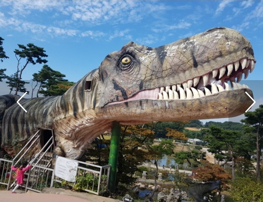 Uma grande estátua de dinossauro com um olho e dentes visíveis, uma estrutura de suporte verde e uma placa.