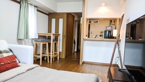 Quarto de hotel com cama, uma pequena mesa com cadeiras, cozinha compacta e banheiro visível por uma porta aberta.