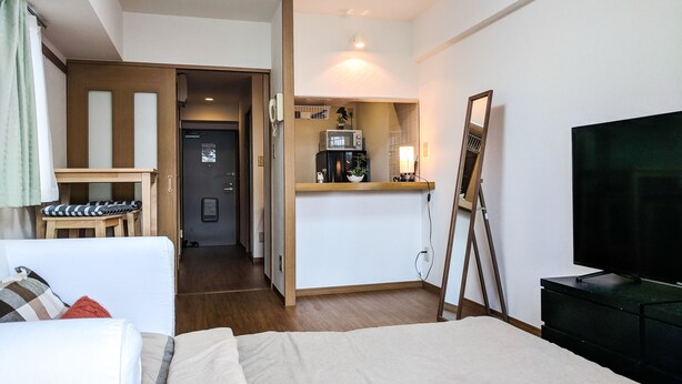 Quarto de hotel com cama, escrivaninha, espelho, televisão e cozinha compacta.