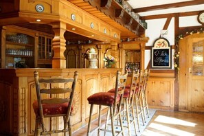 Uma área de bar em madeira com vários bancos e um balcão, apresentando uma lousa com um coração e a palavra 'Willkommen'.