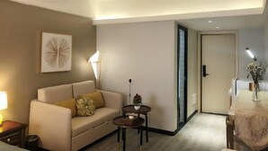 Uma sala de estar moderna com um sofá, uma mesa pequena e uma luminária. Há uma porta que dá para outro quarto.