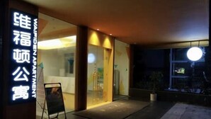 Entrada de um edifício moderno com porta de vidro, uma placa de identificação e interior bem iluminado visível pelo vidro.