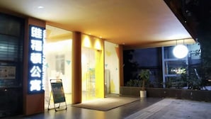 Entrada de um edifício moderno com fachada de vidro, letreiro neon e uma pequena área de estar ao ar livre.