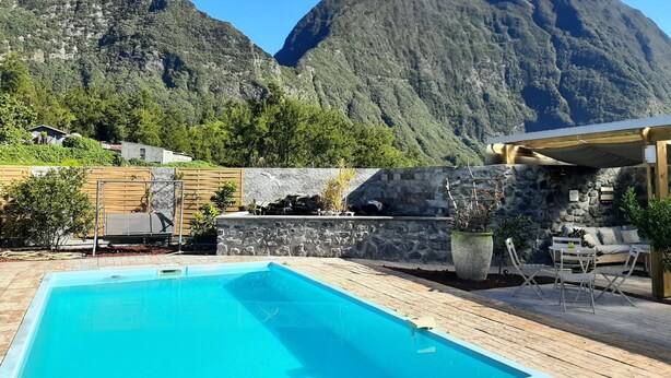 Piscina com montanhas ao fundo, deck de madeira, parede de pedra e área de estar externa.