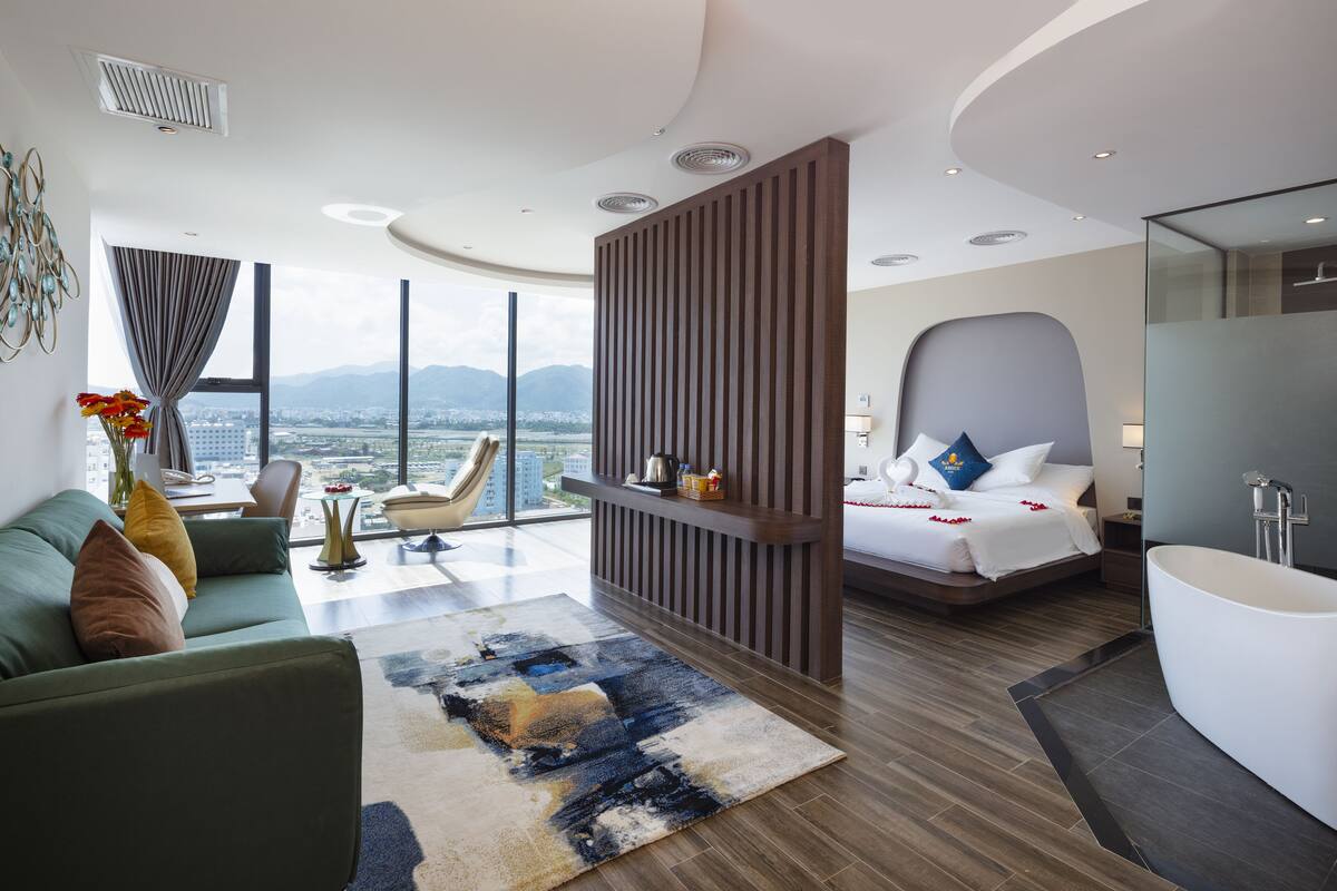 Quarto de hotel moderno com uma cama grande, área de estar com sofá e poltrona, vista da cidade e banheiro privativo com banheira.