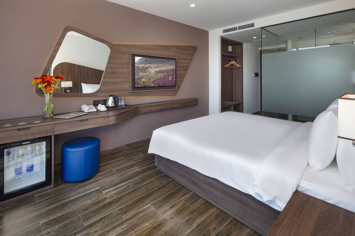 Quarto de hotel moderno com uma cama grande, uma escrivaninha embutida com espelho, um frigorífico e uma lixeira azul.