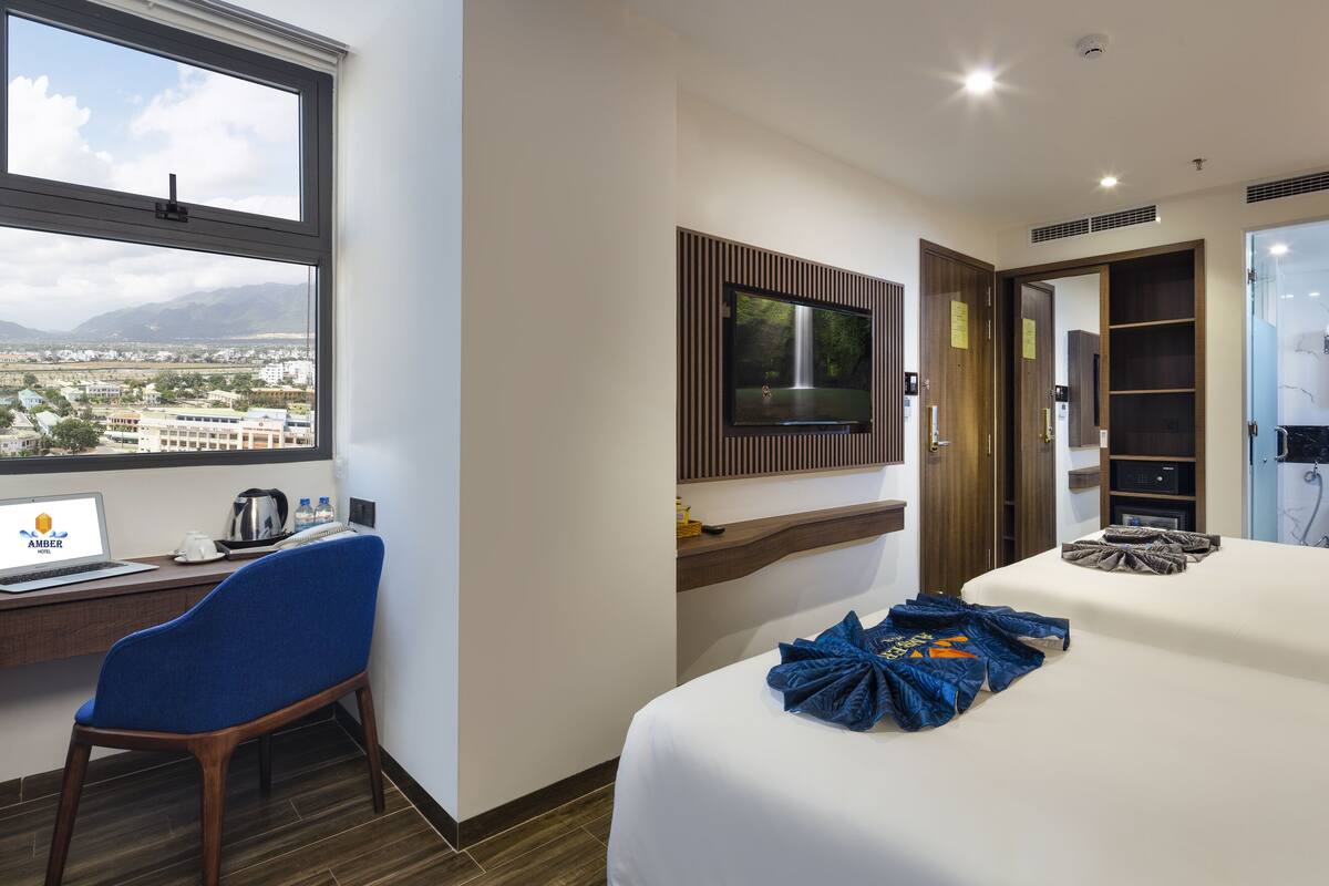 Quarto de hotel com duas camas, uma secretária com um computador portátil, uma televisão e vista para a paisagem urbana.