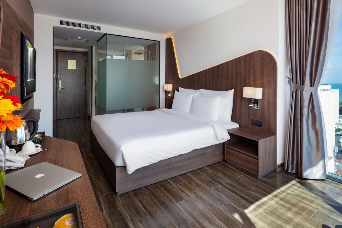 Quarto de hotel moderno com uma cama grande, uma escrivaninha com um laptop, uma mesa de cabeceira com abajur, e vista da cidade pela janela.