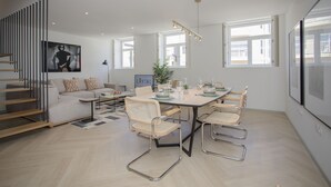 Uma sala de estar moderna com área de jantar, apresentando um sofá, uma mesa de centro, uma mesa de jantar com cadeiras, uma escada e janelas amplas.