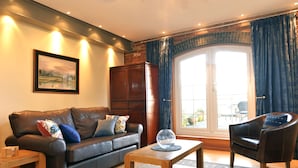 Sala de estar com sofá de couro, mesa de centro de madeira e poltrona de couro escura. Há uma janela grande com cortinas azuis e vista para o exterior.