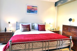 Quarto bem arrumado com estrutura de cama em metal, cobertor vermelho e mesa de cabeceira com abajur.