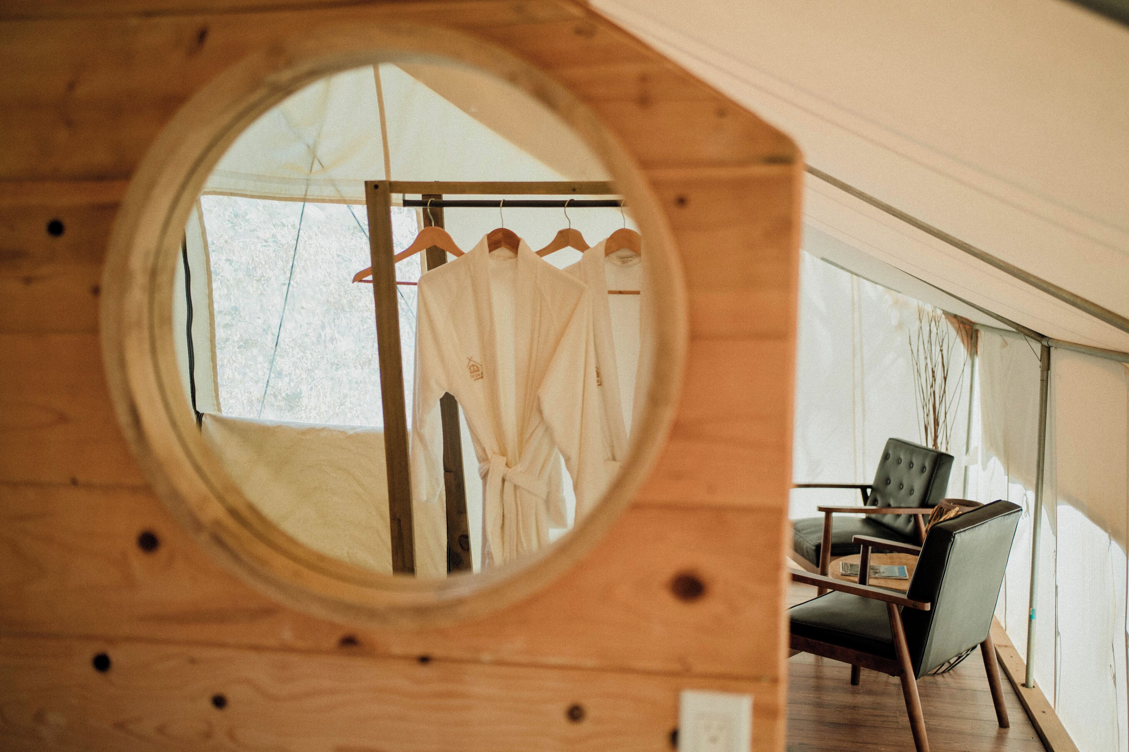 Janela redonda com vista para um quarto que inclui uma cadeira, uma mesa e um cabideiro com roupões.