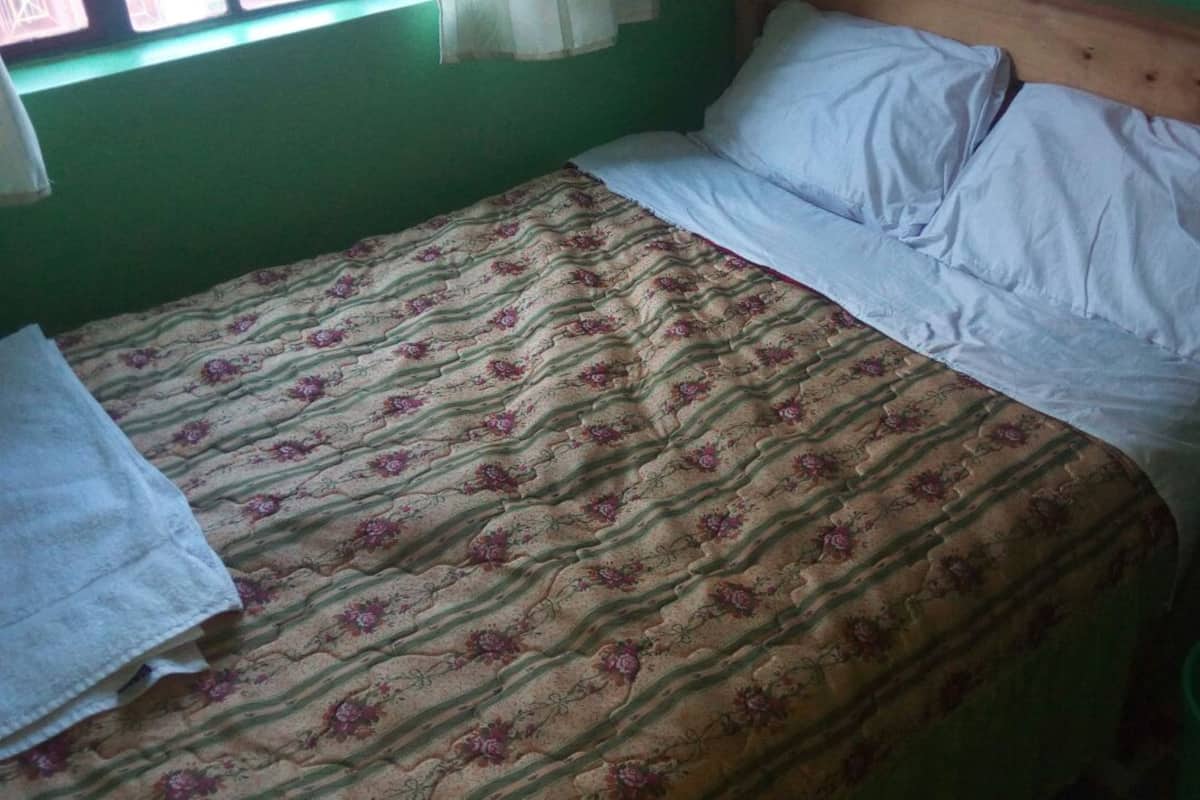 Uma cama de solteiro com colcha estampada em flor, travesseiros brancos e cabeceira de madeira. Uma lixeira verde está visível à direita.