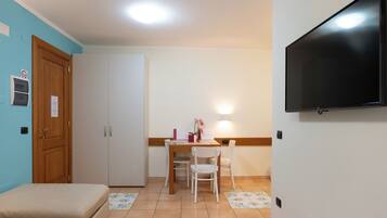 Um quarto com uma cama, uma escrivaninha com cadeiras, uma televisão e uma porta.