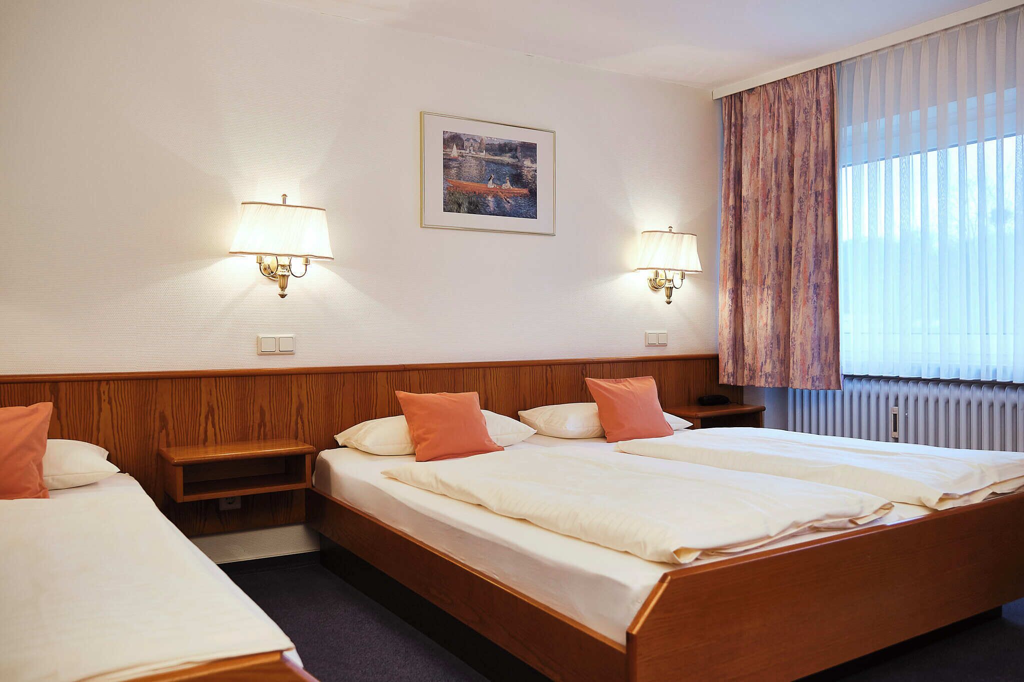 Quarto de hotel com duas camas, cabeceira de madeira, duas mesas de cabeceira, um criado-mudo, luminária na parede, janela com cortinas e um quadro na parede.