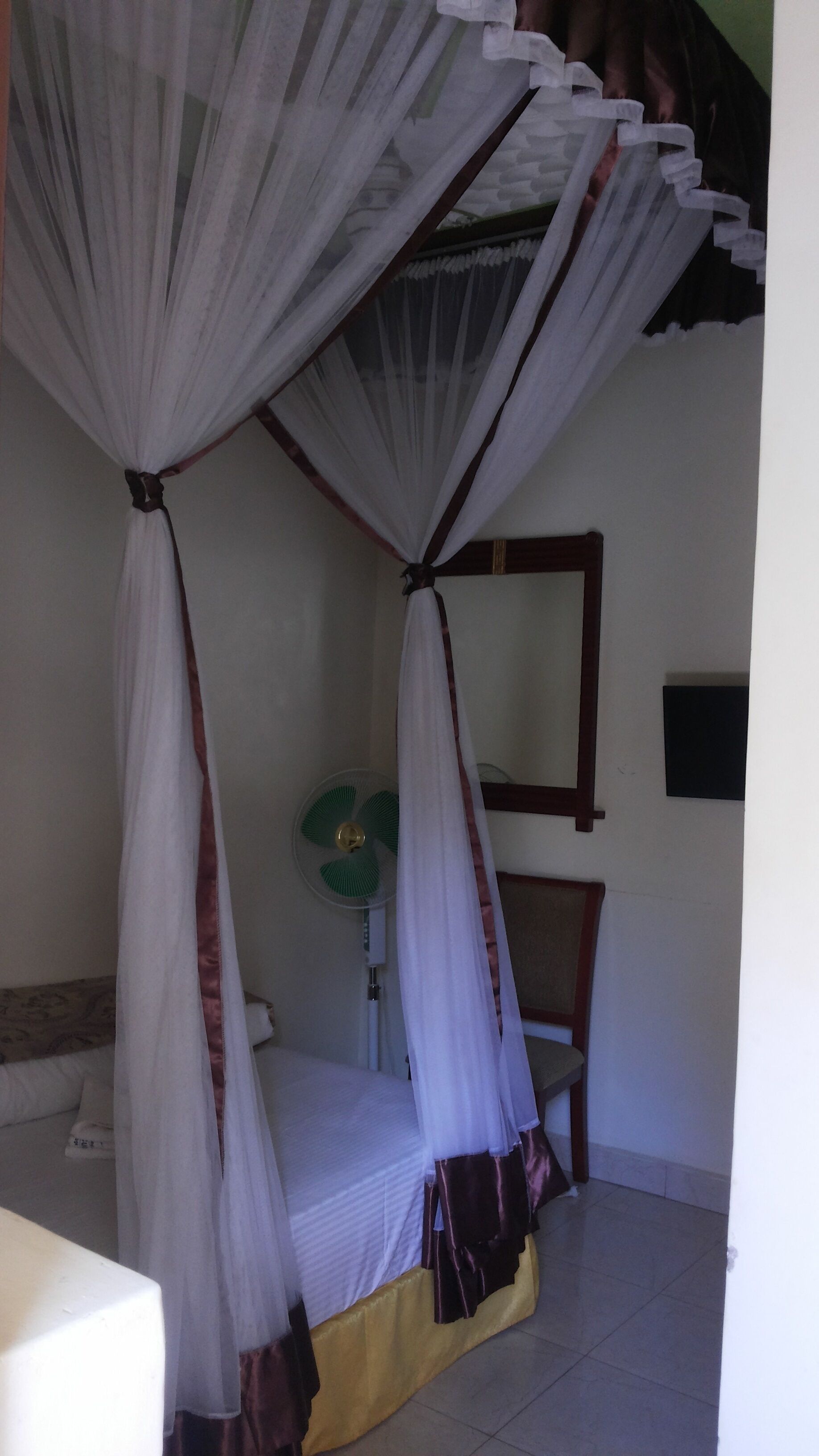 Quarto com cama sob um dossel, mesa de cabeceira, cadeira, espelho fixado na parede e um ventilador.