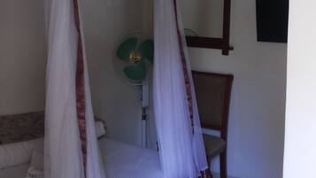 Quarto com cama sob um dossel, mesa de cabeceira, cadeira, espelho fixado na parede e um ventilador.