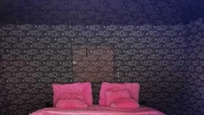 Quarto com colcha rosa, duas almofadas e luminária pendente.