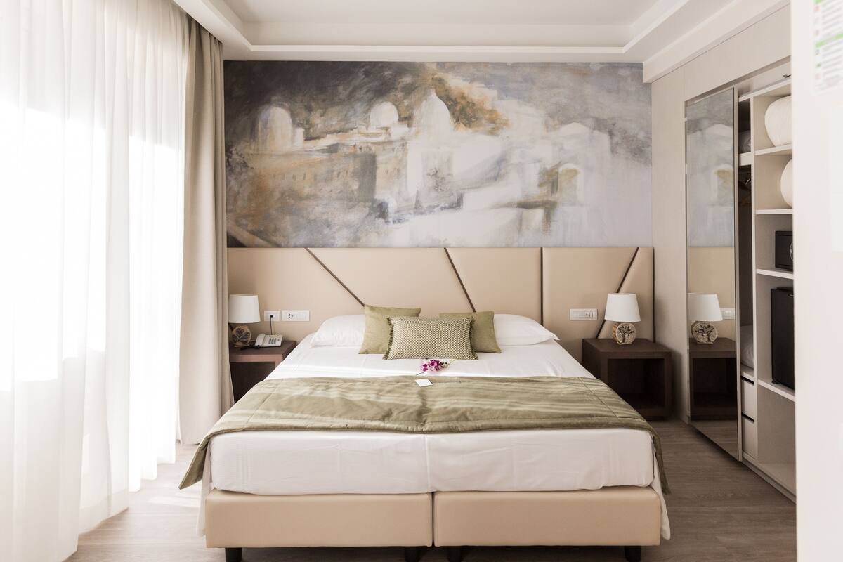 Quarto de hotel com uma cama grande, duas mesas de cabeceira com luminárias, um quadro abstrato grande na parede e um guarda-roupa com espelho.
