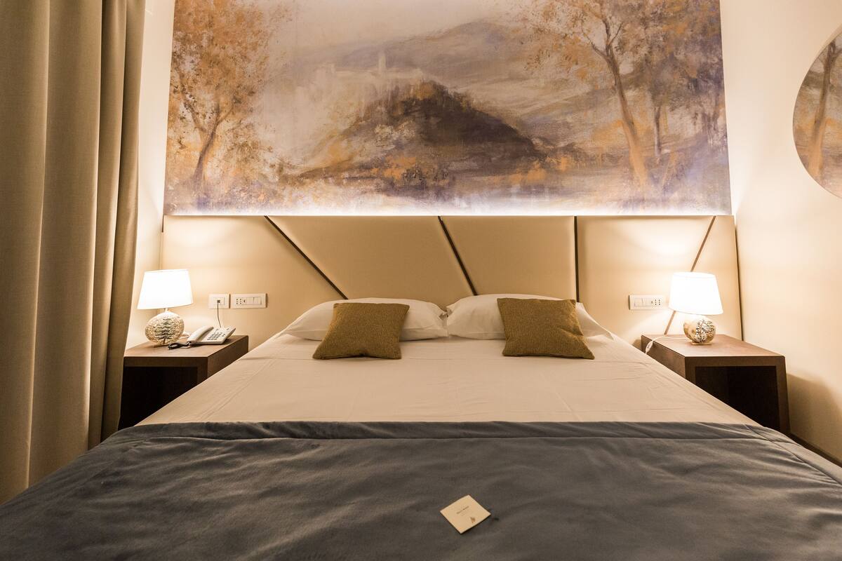 Quarto de hotel com uma cama grande, duas mesas de cabeceira com luminárias e uma pintura sobre a cama.