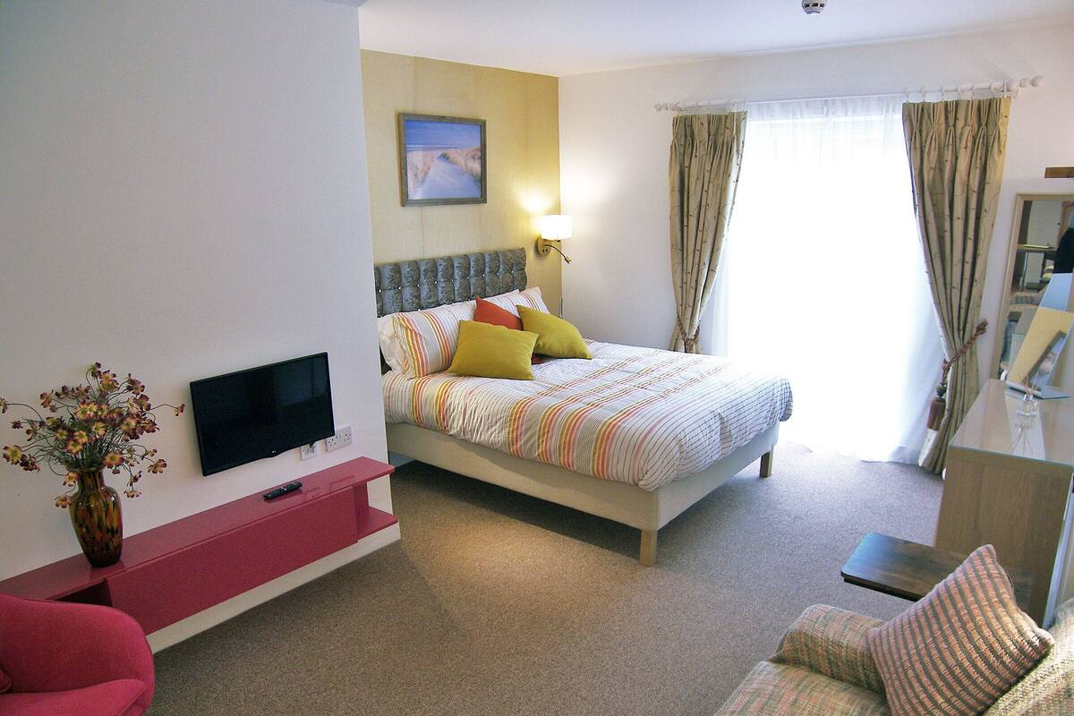 Quarto de hotel com cama, televisão, escrivaninha, cadeira e um vaso com flores.