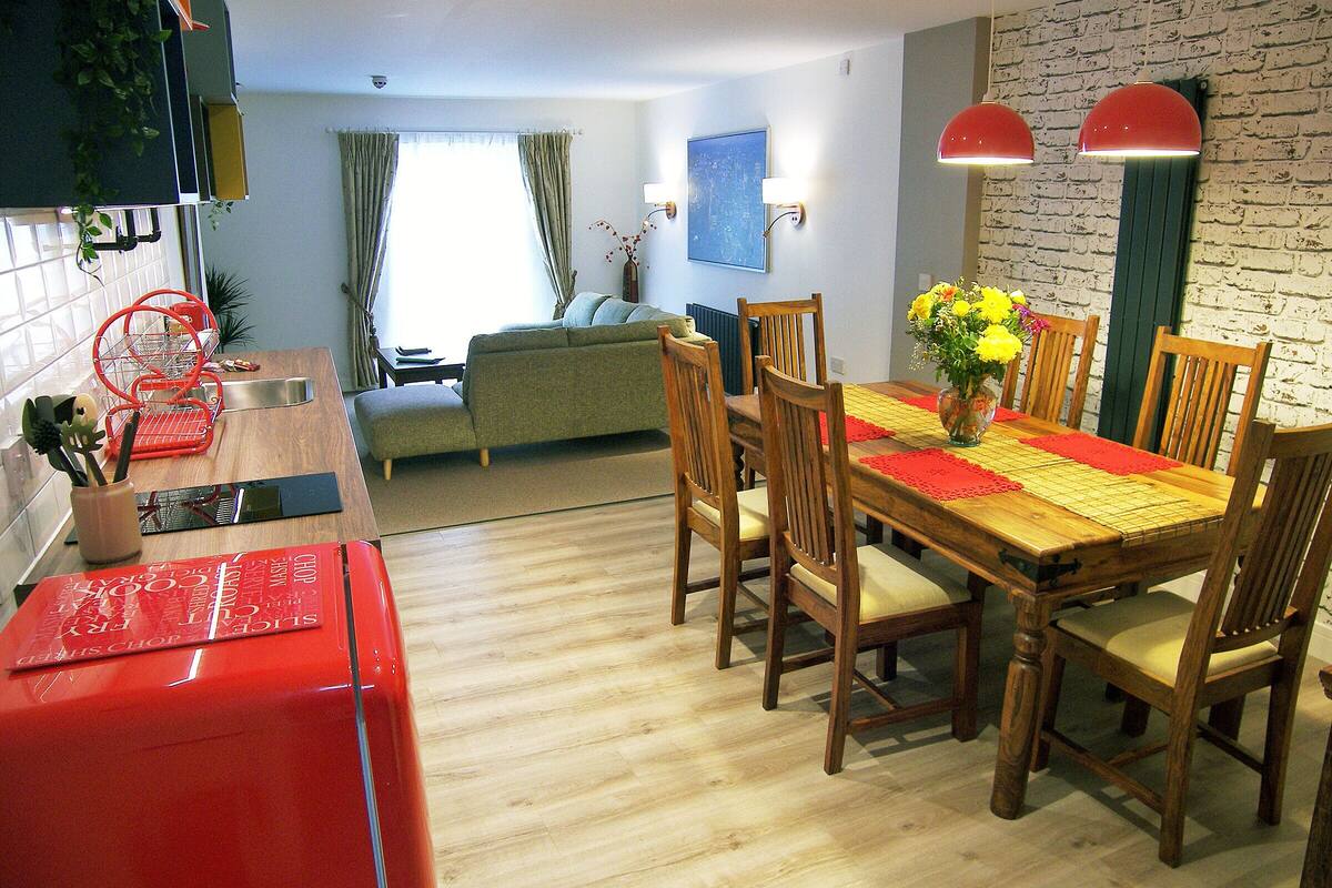 Uma área de refeições com uma mesa de madeira, cadeiras e um jogo americano vermelho. Há um sofá e uma janela com cortinas ao fundo.