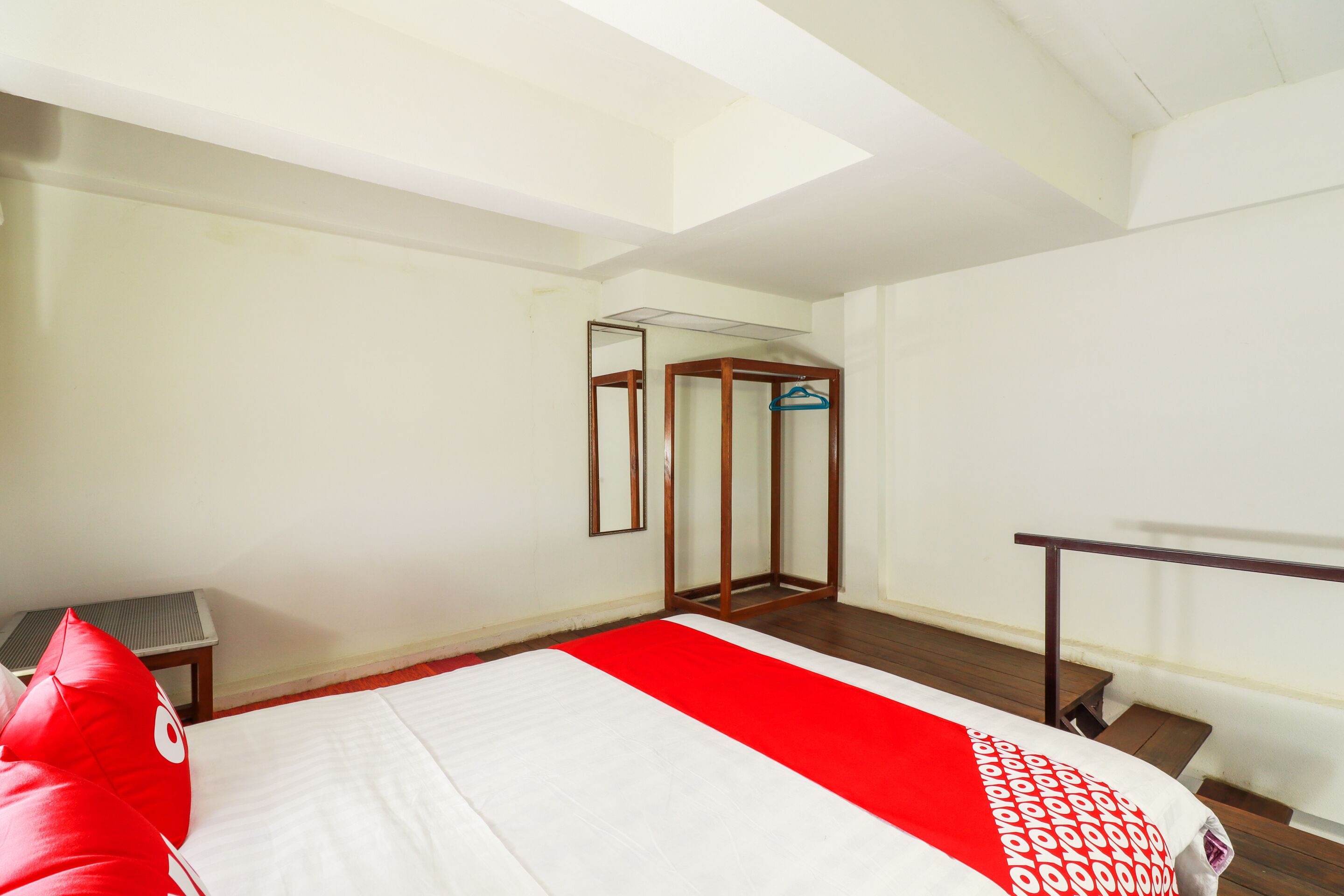 Quarto de hotel com cama, colcha vermelha e branca, mesa de cabeceira de madeira, espelho e vista para o corredor.